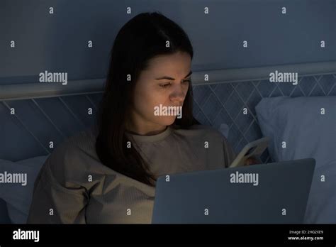Woman Using Laptop Computer And Surfing Social Media On Smartphone In Bed Before Sleep At Night