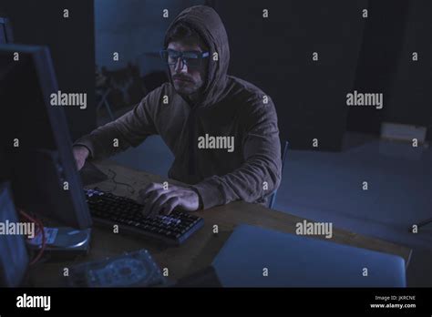 Serious Computer Hacker Wearing Hooded Shirt Using Desktop Computer At Table In Abandoned Room