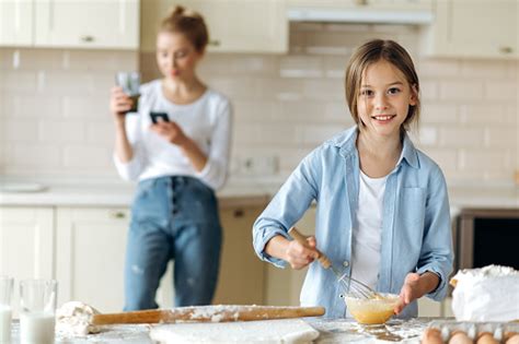 엄마의 작은 도우미 귀여운 백인 어린 소녀는 파이를 요리하는 법을 배운다 계란을 친다 카메라를보고 미소 엄마는 전화를 사용하여 배경에 서 2명에 대한 스톡 사진 및 기타