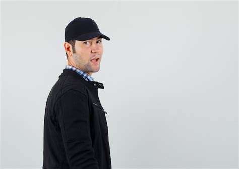 Молодой человек смотрит в камеру через плечо в рубашке куртке кепке и выглядит уверенно