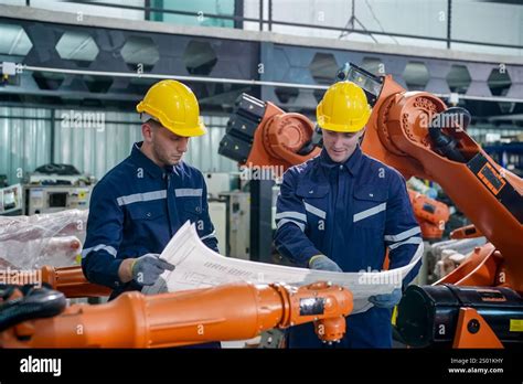 Two Engineers Wearing Hard Hats Examine Blueprints For A Robotic System Within A Contemporary