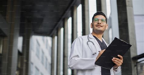 클립보드에 Rx 처방전을 쓰는 잠겨 있는 전문 남성 아랍 의사의 초상화를 닫습니다 남성 의사는 현대 클리닉의 야외에 서서 의료 양식을 작성하고 있습니다 복사 공간 사진