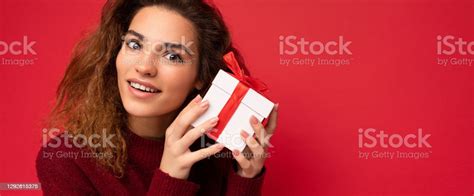 매력적인 긍정적 인 미소 젊은 갈색 머리 곱슬 여자의 클로즈업 사진 빨간 배경 벽에 고립 된 빨간 스웨터를 입고 선물 상자를 들고 카메라를보고 꿈꾸는 귀여운에 대한 스톡