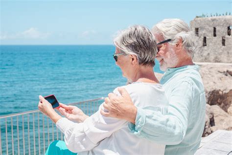 바다 휴가를 즐기는 선글라스를 착용 한 백인 노인 부부의 뒷모습 앉아서 휴대 전화를 사용하여 포옹 노인을 웃고 물 위의 푸른 지평선 공간 복사 2명에 대한 스톡 사진 및