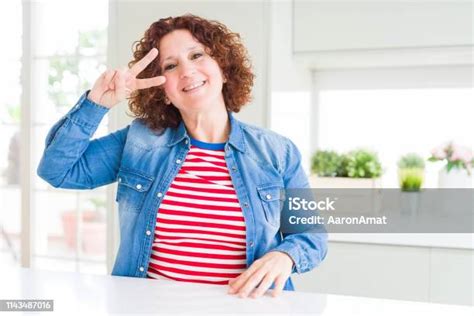 곱슬 머리를 가진 중간 나이 고위 여자는 얼굴에 손가락으로 평화 기호를 하 고 집에서 데님 재킷을 입고 쾌활 한 표시 승리를 웃는 가정 생활에 대한 스톡 사진 및 기타