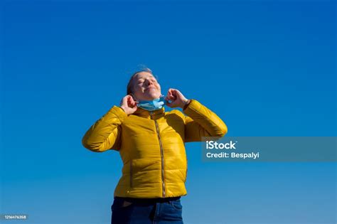 소녀는 유쾌한 표정으로 얼굴에서 의료 용 마스크를 제거하고 격리 중에 신선한 공기를 흡입 복사 공간이있는 푸른 하늘에 노란색 재킷을 입은 사람 개념에 대한 스톡 사진 및