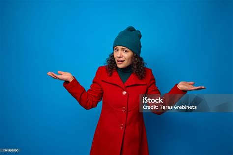 깜짝 놀란 젊은 곱슬 머리 갈색 갈색 머리 여자 에 빨간 코트와 녹색 모자 포즈 에 푸른 배경에 대 한 포즈 그녀의 손 손바닥에 상상의 복사 공간을 들고 카메라를 보고