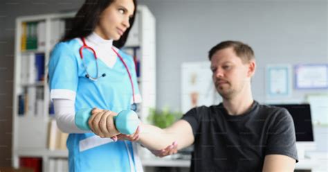 의사는 병원에서 손을 느낍니다 남자는 의사 진료 시간에 덤벨로 팔을 앞으로 뻗은 채 의자에 앉습니다 의사복을 입고 빨간 청진기를 쓴 여성이 방문객의 손 골절 여부를