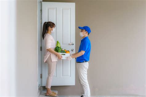 얼굴 마스크와 장갑을 착용한 아시아 여성 의상에는 집 앞에서 배달하는 남자로부터 음식 과일 채소 음료의 식료품 상자를 받습니다 20 24세에 대한 스톡 사진 및 기타 이미지