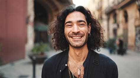 긴 곱슬 머리와 그루터기를 가진 젊은 매력적인 이탈리아 남자는 카메라를 들여다 보며 오래된 건물 배경을 미소 짓습니다 귀에 귀걸이와 많은 체인을 가진 세련된 남자 감정에