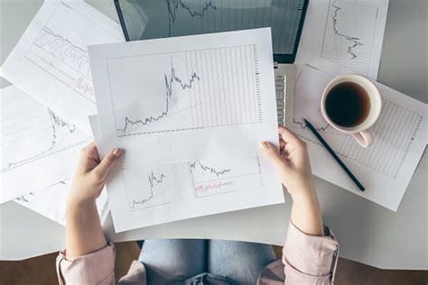 맨 위 보기 커피 한 잔을 들고 테이블에 앉아 종이에 인쇄 된 그래픽과 차트가있는 노트북에서 일하는 젊은 비즈니스 여성 거래자에