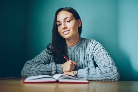 대학 인테리어의 나무 책상에서 공부하는 노트북에서 할 수있는 카메라를 쓰고 있는 갈색 머리를 가진 지능형 젊은 여성의 절반 길이
