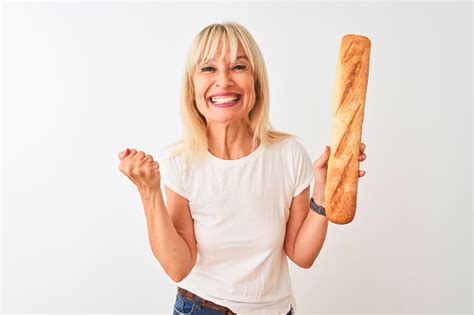 고립된 흰색 배경 위에 빵을 들고 있는 중년 제빵사 여성은 자랑스럽고 승리와 성공을 축하하며 매우 흥분하고 환호하는 감정을