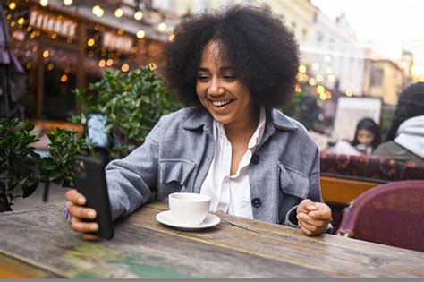 아프로 머리를 가진 매력적인 젊은 행복한 아프리카 계 미국인 여성의 거리 스타일의 초상화는 보도 카페에서 야외에서 스마트 폰과