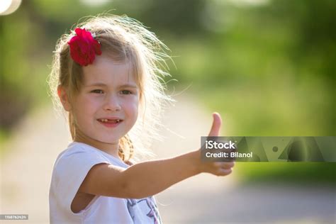 흐릿한 밝은 녹색 여름 배경에 카메라에 엄지 손가락 제스처를 보여주는 머리에 빨간 장미와 흰색 티셔츠에 귀여운 작은 금발 심각한 소녀의 흐릿한 초상화 어린 시절 개념의