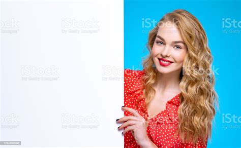 젊은 여성과 큰 흰색 배너 아름다운 웃는 소녀가 빌보드 옆에 서 있습니다 유럽의 쾌활한 여성이 광고 배너 옆에 서 있습니다 금발 소녀와 종이의 빈 광고 시트 20 24세에