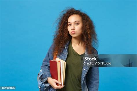 데님 옷에 젊은 아프리카계 미국인 소녀 사춘기 학생 배낭은 파란색 벽 배경 스튜디오 초상화에 고립 된 책을 잡아 고등학교 대학