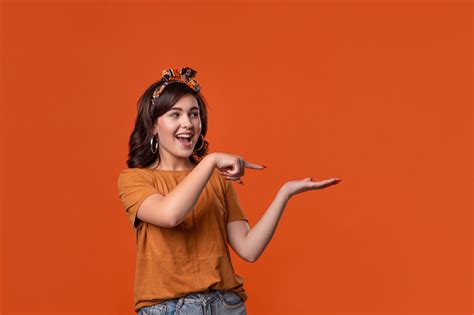 티셔츠와 머리띠를 입고 오렌지 색 배경 위에 고립 된 제품 배치를위한 손바닥을 들고있는 아름다운 미소 갈색 머리 여성 광고를 위한 장소 T 셔츠에 대한 스톡 사진 및 기타