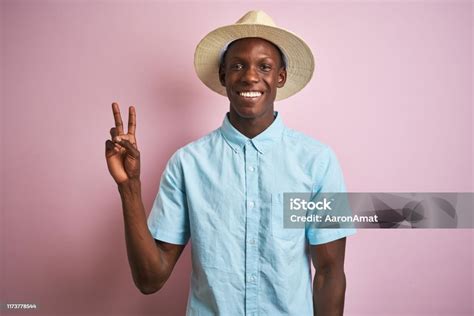 파란색 셔츠와 여름 모자를 쓰고 고립된 분홍색 배경 위에 행복한 얼굴이 승리의 표지판을 들고 미소짓고 있습니다 2번 긍정적인 감정 표현에 대한 스톡 사진 및 기타 이미지