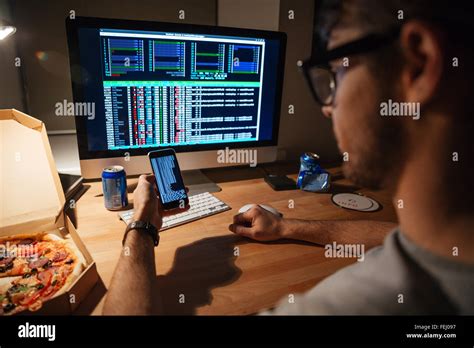 Closeup Of Serious Young Developer In Glasses Using Computer And Smartphone For Coding At Home