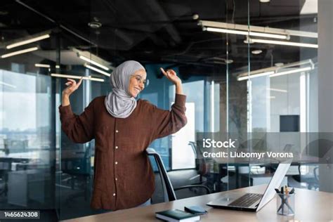 직장에서 춤을 추는 즐겁고 성공적인 사업가 직장에서 승리와 성공적인 성취를 축하하는 히잡을 쓴 무슬림 여성 사무실 내부에서 좋은 분위기를 가지고 있는 아랍인 노트북으로