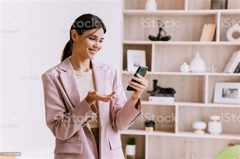 분홍색 정장을 입은 쾌활한 아시아 갈색 머리 사업가가 전화를 들고 책장에 기대어 집에서 화상 통화를 합니다 좋은 소식에 놀란