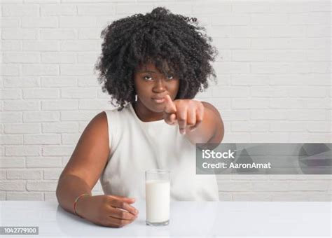 카메라 하 고 당신에 게 손 기호 긍정적이 고 자신감 제스처 앞에서 손가락으로 가리키는 우유 한 잔을 마시는 테이블에 앉아 젊은 아프리카계 미국인 여자 가리키기에 대한 스톡