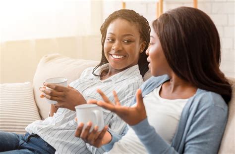 Two Black Girls Sitting Together On Sofa Chatting And Drinking Tea