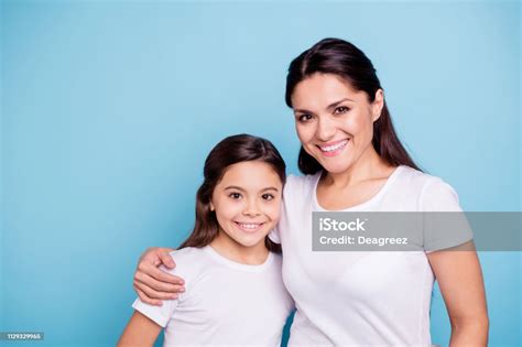 사진 놀라운 두 사람 갈색 머리 엄마 작은 작은 딸 서 포옹 사랑 스러운 자유 시간을 꽤 가까이 밝은 파란색 배경에 고립 된 흰색 티셔츠를 입고 즐 거 워 라틴 아메리카