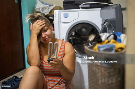 화가 난 여성이 발열체를 손에 들고 고장난 세탁기 옆에 앉아 있다 더러운 빨래 바구니를 든 한 여자가 집 세탁기 근처에서 슬퍼하고 있다 깨짐에 대한 스톡 사진 및 기타