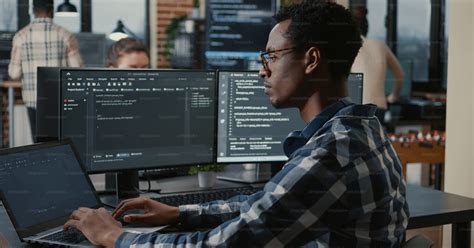 집중된 프로그래머는 노트북에 타이핑하는 동안 안경을 고정하고 책상에 앉아 여러 화면으로 코드를 구문 분석합니다 코더는 백그라운드에서 빅 데이터를 코딩하는 프로그래머 팀과