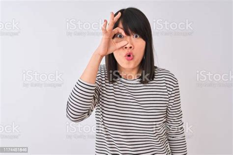 고립 된 흰색 배경 위에 검은 줄무늬 티셔츠를 입은 젊은 아름다운 중국 여성은 손가락을 통해 눈을 보고 놀란 얼굴로 충격을 받은 확인 제스처를하고 있습니다 믿을 수 없는