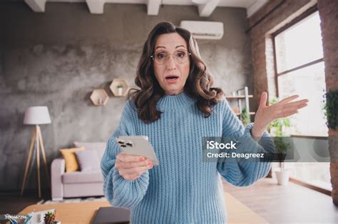현대적인 장식과 일광이 들어오는 아늑한 거실에서 스마트 폰을 보는 놀란 노인 여성 65 69세에 대한 스톡 사진 및 기타 이미지 65 69세 Brand Name