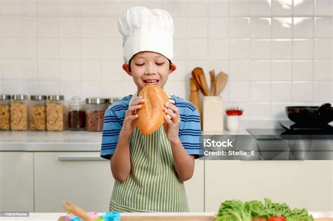 큰 빵 덩어리를 먹는 아이의 초상화 앞치마와 요리사 모자를 쓰고 부엌에서 갓 구운 수제 빵을 먹는 행복한 아시아 소년 귀여운