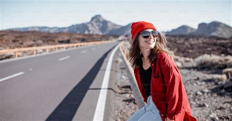 화창한 날 화산 계곡 한가운데에 있는 도로 울타리에 앉아 빨간 옷을 세련되게 입은 젊은 여성의 라이프스타일 초상화 평온한 라이프 스타일과 여행 컨셉 사진
