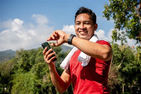 A Happy Asian Man In Sportswear Checks His Calorie Count On His Smartwatch While Jogging In A