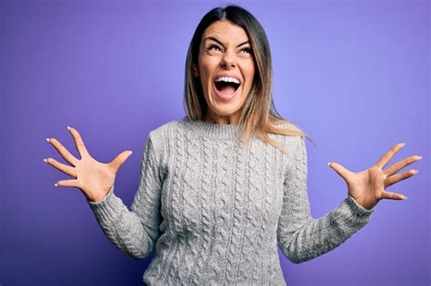 고립 된 보라색 배경 위에 서있는 캐주얼 스웨터를 입은 젊은 아름다운 여성은 공격적인 표정과 팔을 들고 미친 소리와 고함을 지으며 좌절 개념입니다 갈색 머리에 대한 스톡