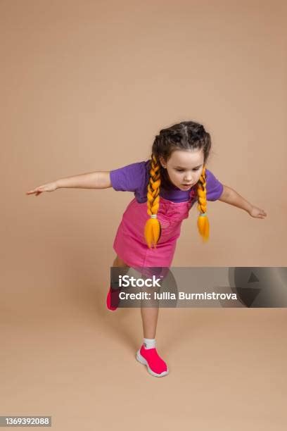 분홍색 점프수트와 베이지색 배경에 보라색 티셔츠를 입고 아래를 내려다보며 옆으로 손을 대고 한쪽 다리에 서 있는 노란 카네칼론 머리띠를 가진 장난스러운 어린 소녀의 초상화