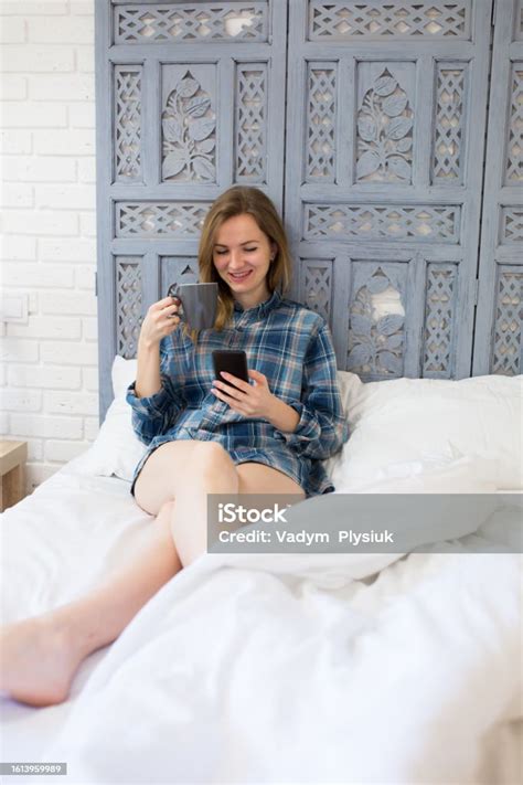 아름 다운 여자 흰 침대 시트에 앉아 있다 남자의 체크 무늬 셔츠에 젊은 여성이 커피를 마시는 이며 흰색 베개에 소셜 네트워크에서 검색 손에 검은 스마트폰으로 아침입니다