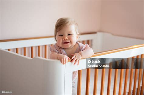생후 6개월 된 귀여운 백인 여자 아이가 유아용 침대에 서서 카메라를 보고 있는 모습 6 11 개월에 대한 스톡 사진 및 기타