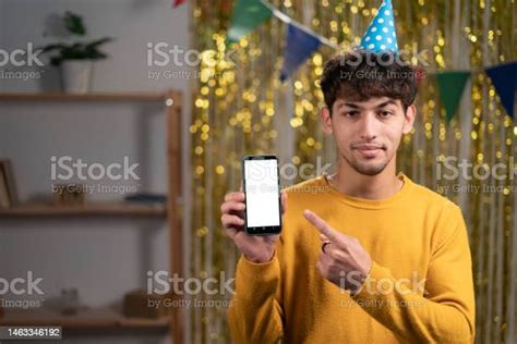 흰색 화면이 비어 있는 휴대폰을 손가락으로 가리키는 축제 모자를 쓴 청년의 모형 이미지 집에서 생일 파티 축하 공란에 대한 스톡 사진 및 기타 이미지 Istock