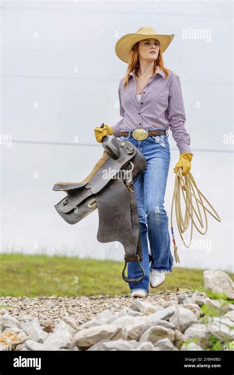 A Beautiful Redhead Model Poses In A Country Setting Wearing Cowgirl Attire And Holding A Saddle