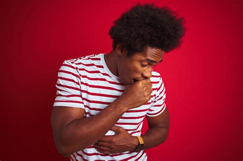 고립 된 빨간 배경 위에 줄무늬 티셔츠를 입고 아프리카 계 미국인 남성이 감기 또는 기관지염의 증상으로 기분이 좋지 않고 기침합니다 헬스케어 개념 T 셔츠에 대한 스톡 사진