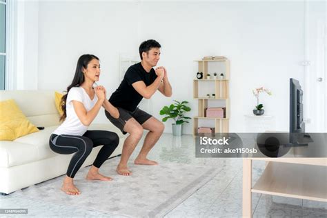 젊은 아시아 부부는 함께 스쿼트 훈련을하고 집에서 Tv를 찾고 남자와 여자는 함께 거실에 서 운동 파트너와 피트니스 운동을 수행하는 쌍에 맞는 운동에 대한 스톡 사진 및