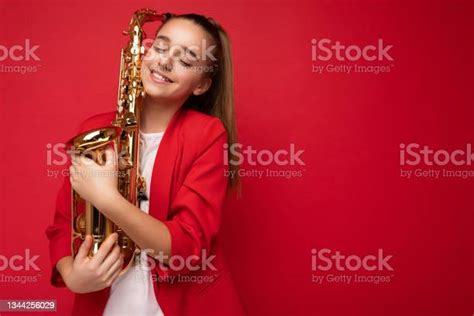 색소폰을 들고 눈을 감고 꿈꾸는 빨간 배경 벽에 고립 된 트렌디 한 빨간 재킷을 입은 꽤 귀여운 긍정적 인 미소 브루넷 어린 소녀의 샷 색소폰에 대한 스톡 사진 및 기타