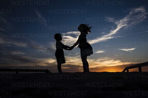 Silhouette Sisters Holding Hands While Standing On Field Against Sky