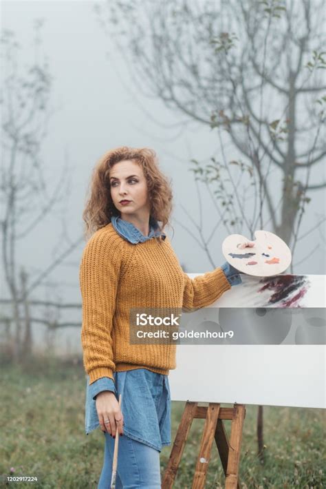 야외에서 그림을 그리는 젊은 여성 예술가 그녀는 캔버스와 그림 앞에 있습니다 그녀는 오일 페인트 아티스트 브러쉬 캔버스 및 팔레트를 보유하고 있습니다 곱슬 머리를 가진 농축