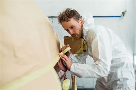 자동 화가 마스킹 차 체에 테이프와 종이 다시 그리기 전에 흰색 보호복을 입은 정비사가 차고에서 작동합니다 25 29세에 대한