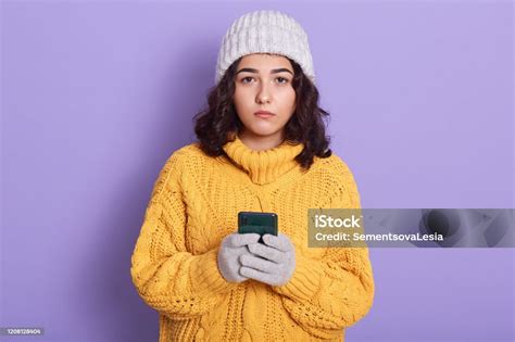 카메라를 직접 보고 라일락 배경 위에 고립 된 서 따뜻한 옷을 입고 양손에 스마트 폰을 들고 화가 실망 갈색 머리의 수평 실내 샷 기술 개념 겨울에 대한 스톡 사진 및 기타