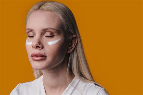 노란색 배경 위에 고립된 얼굴에 보습 크림을 바르는 매력적인 금발 장발 여성의 초상화 젊은 여자 얼굴 초상화 테라피 트리트먼트 뷰티 스킨 케어 미녀는 피부 관리에 직면합니다